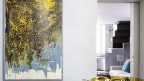 Decoração moderna com um quadro abstrato grande, uma tigela de vidro sobre uma mesa, e vista para uma sala de estar com escada.