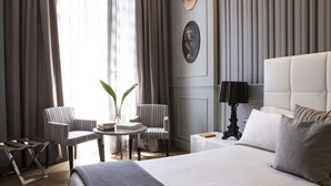 Quarto de hotel com uma cama grande, duas cadeiras, uma mesa pequena com livros, um abajur e cortinas.