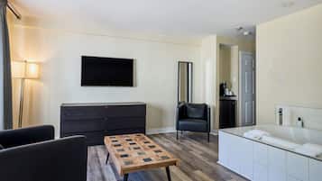 Quarto de hotel moderno com televisão de tela plana, sofá, pequena mesa de madeira, cadeira, espelho grande, banheira e máquina de café.