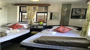 Quarto de hotel com duas camas de solteiro, uma mesa com telefone, uma mesinha com um vaso de flores e um ar condicionado fixado na parede.