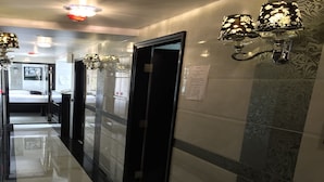 Corredor de hotel com piso refletivo, luminárias fixadas na parede e uma porta para um quarto.