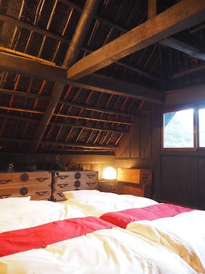 Quarto rústico com teto inclinado, vigas de madeira, cama com roupa de cama branca e vermelha, cômoda de madeira e uma janela com vista.