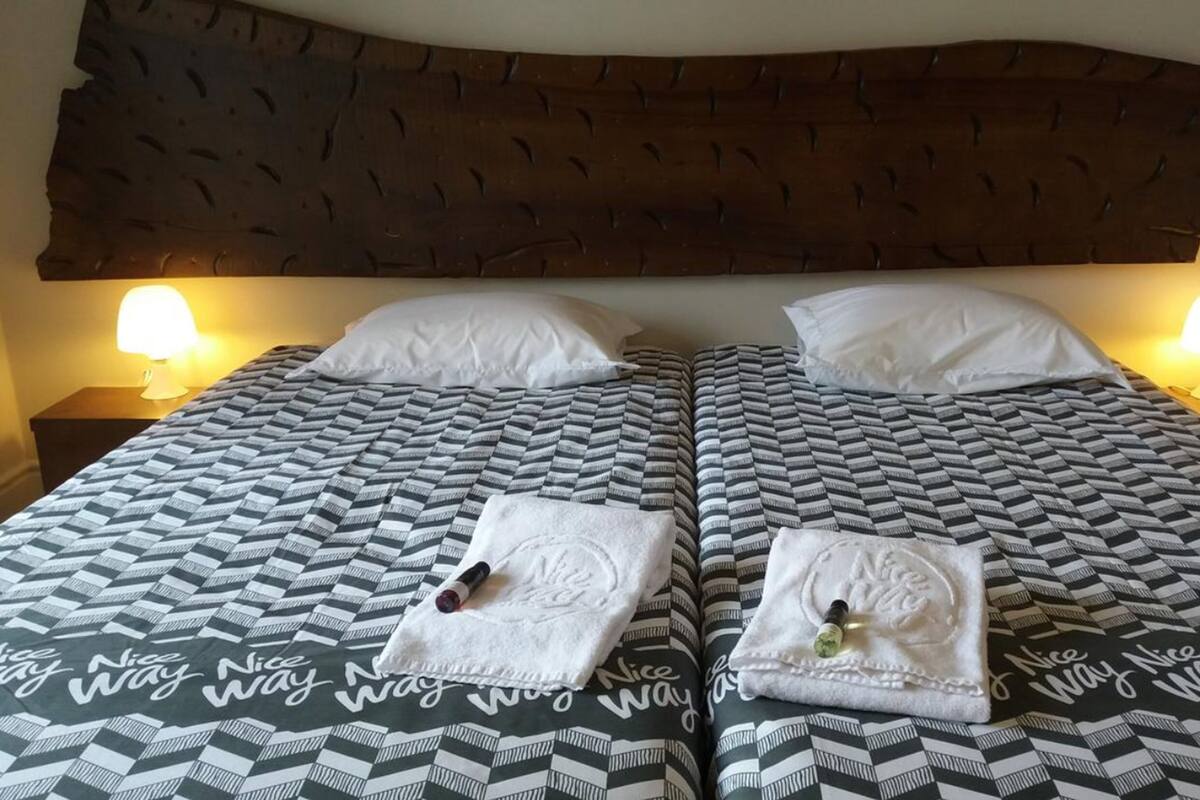 Cama de casal com colcha estampada em padrão geométrico e duas almofadas. Duas luminárias de cabeceira em cada lado da cama.