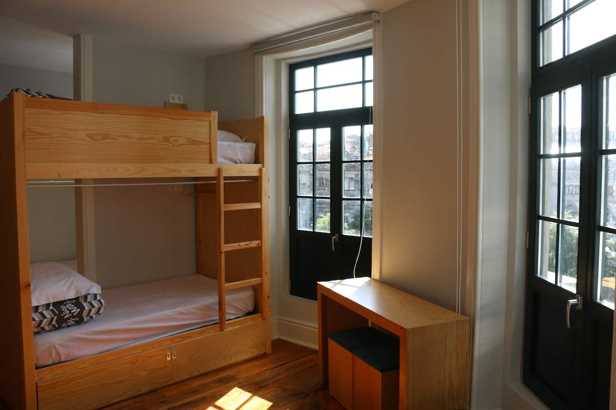 Quarto com beliche, escrivaninha e janela com vista para prédios.