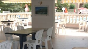 Uma área externa com assentos de cadeiras e mesas de plástico branco, piscina ao fundo e uma grade com bandeiras.