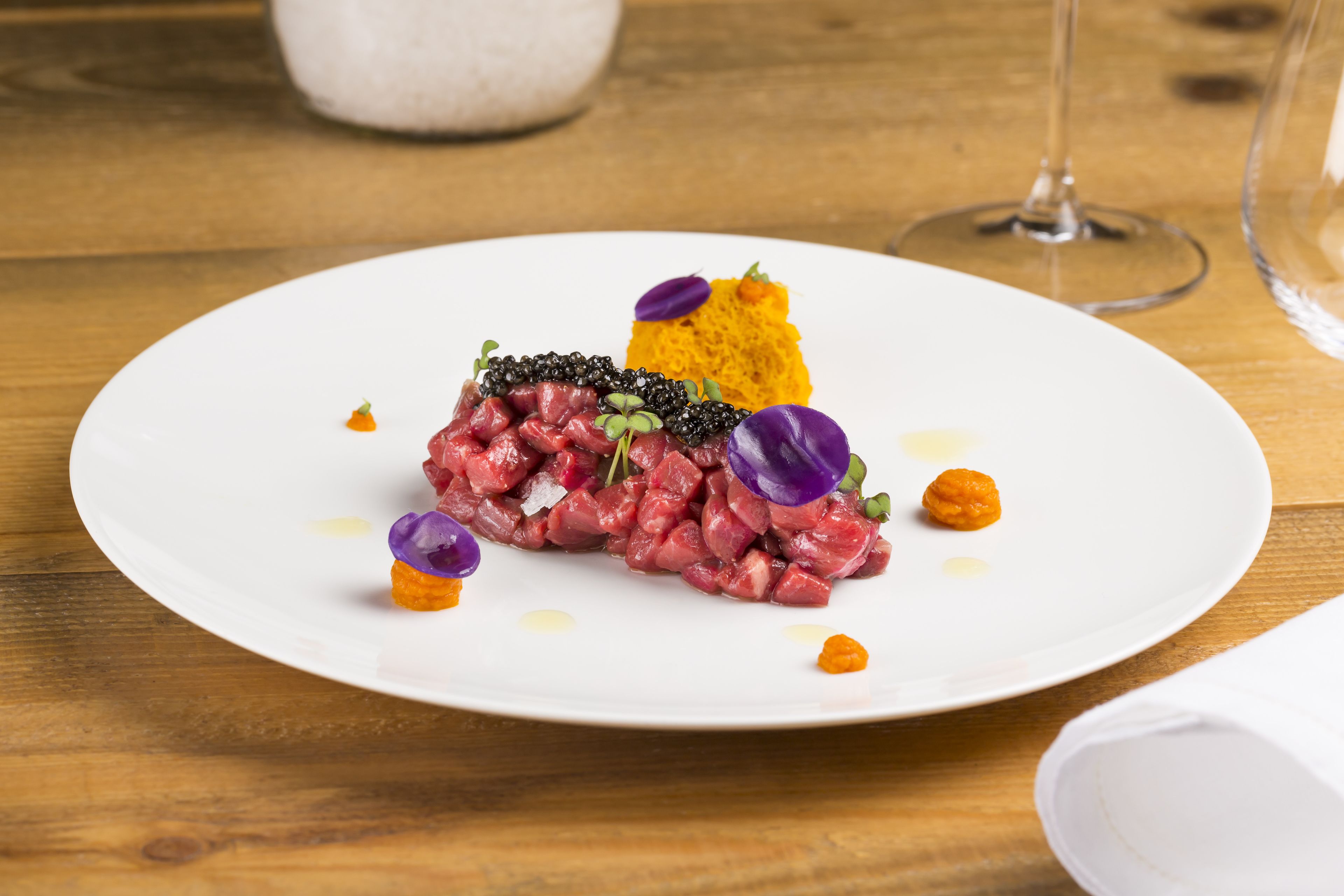 Um prato branco com uma porção de carne crua, decorada com flores roxas e caviar preto, sobre uma mesa de madeira.