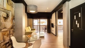 Quarto de hotel moderno com mesa de jantar de vidro, cadeiras brancas, um sofá e uma grande pintura abstrata na parede.