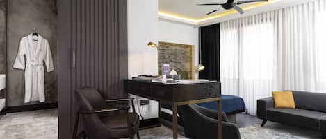 Quarto de hotel com escrivaninha, duas cadeiras, sofá, roupão e uma janela grande com cortinas.