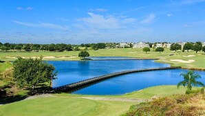Campo de golfe com lago, ponte e um conjunto de edifícios ao longe.