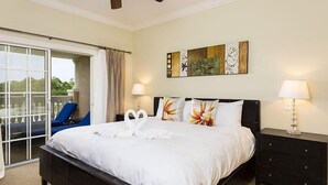Quarto de hotel com uma cama grande, duas luminárias de cabeceira, um ventilador de teto e vista para uma varanda com cadeiras azuis através de uma porta de vidro deslizante.