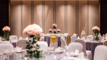 Uma sala de banquetes com mesas redondas preparadas para um evento formal, com cadeiras brancas e fitas amarelas, centros de mesa florais e um bolo em camadas sobre uma mesa.