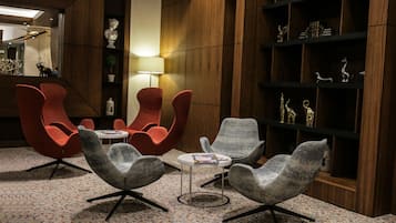 Uma área de estar moderna com uma mistura de cadeiras vermelhas e cinzentas, uma mesa redonda pequena e uma estante com objetos decorativos.