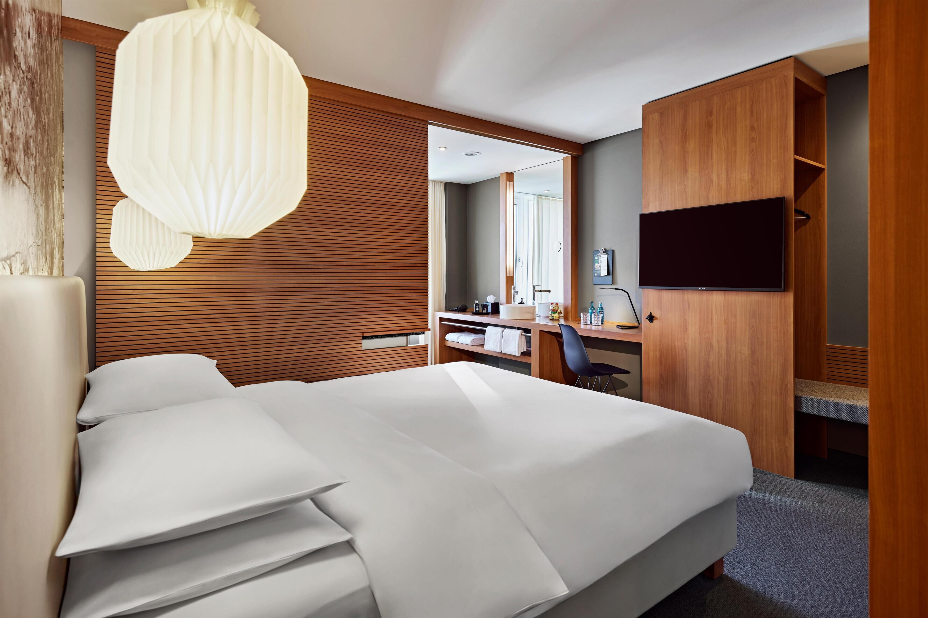 Quarto de hotel moderno com uma cama grande, uma escrivaninha, uma cadeira, uma televisão e um espelho.