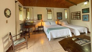 Quarto com vigas de madeira no teto, duas camas com roupa de cama branca, uma escrivaninha com uma cadeira, uma mesa com um controle remoto e obras de arte nas paredes.