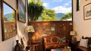 Quarto aconchegante com um banco de madeira, uma mesa com um livro e um vaso decorativo, e vista para montanhas através de uma janela ampla.