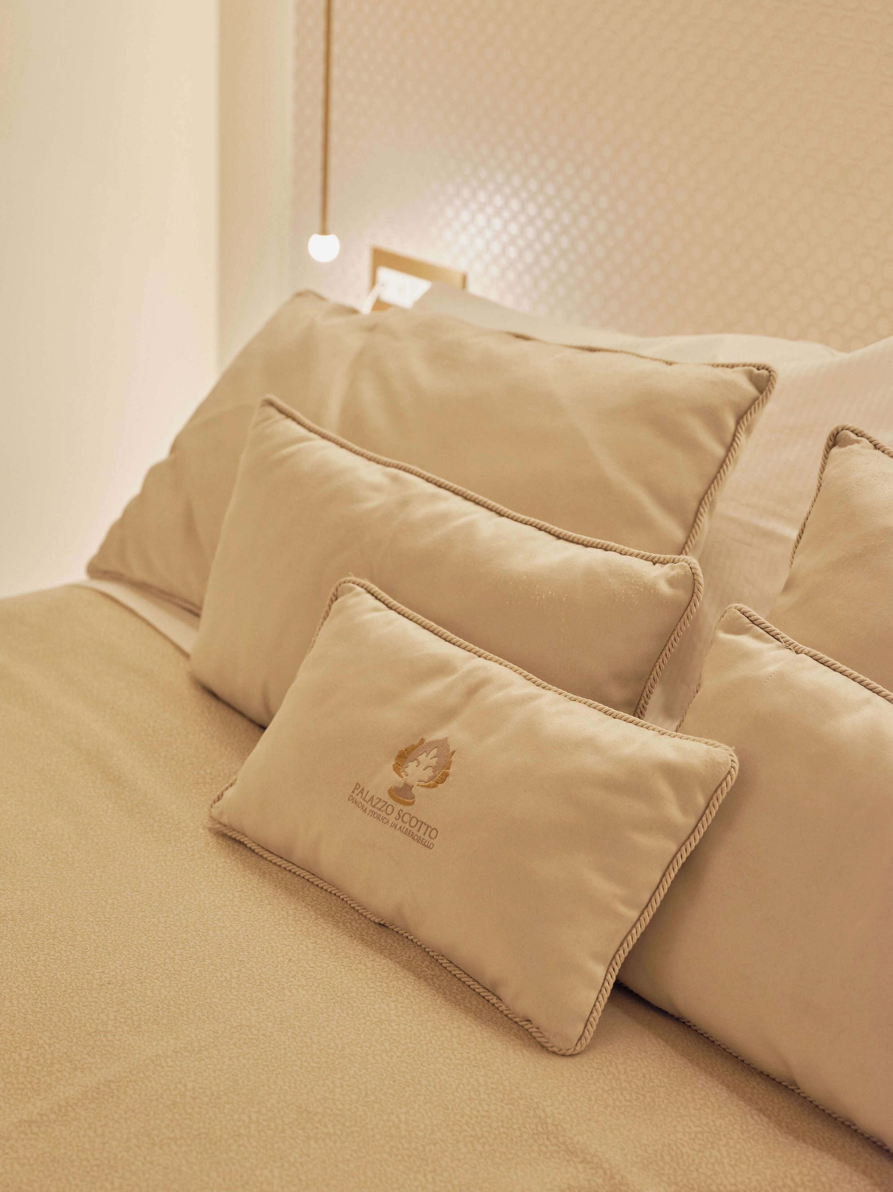 Uma cama com roupa de cama e almofadas bege, incluindo uma almofada menor com um logotipo.