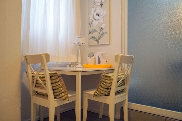 Uma sala de jantar com duas cadeiras brancas, uma mesa, um abajur, uma cafeteira e um quadro na parede.