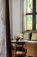 Um sofá de vime com uma mesa de vidro na frente, um vaso com flores e vista para o verde lá fora.