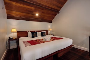 Quarto de hotel com teto de madeira, uma cama grande com roupa de cama branca, duas luminárias de cabeceira e um cobertor decorativo vermelho.