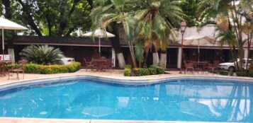 Piscina com espreguiçadeiras e guarda-sóis, cercada por palmeiras e um edifício com área de estar externa.