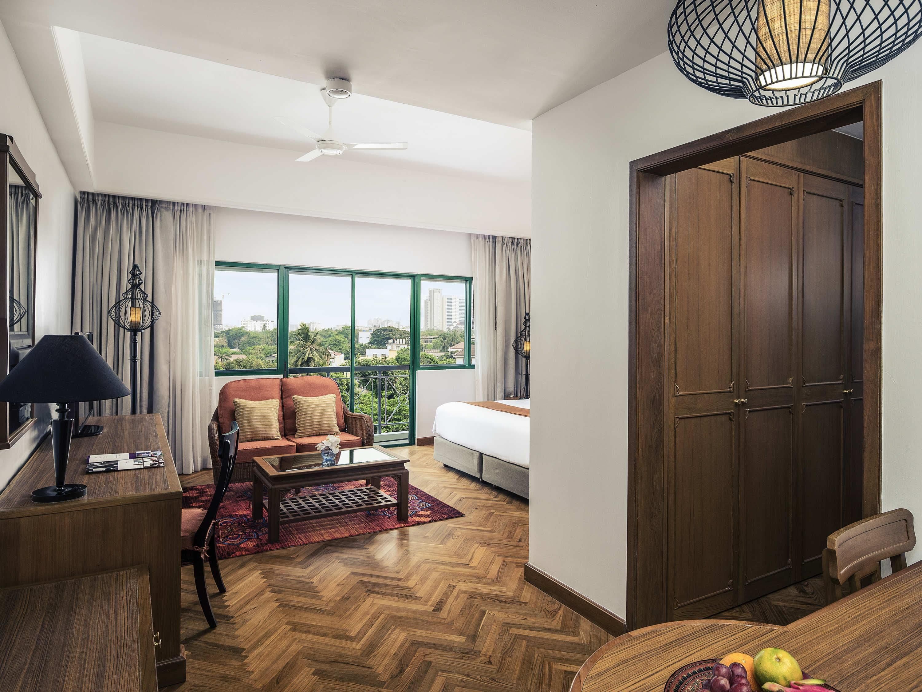 Quarto de hotel com uma janela grande, ventoinha no teto, guarda-roupa de madeira, sofá, mesa de centro e mesa de jantar com frutas.