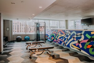 Uma academia com equipamentos de exercícios, um mural colorido e vista da cidade por grandes janelas.