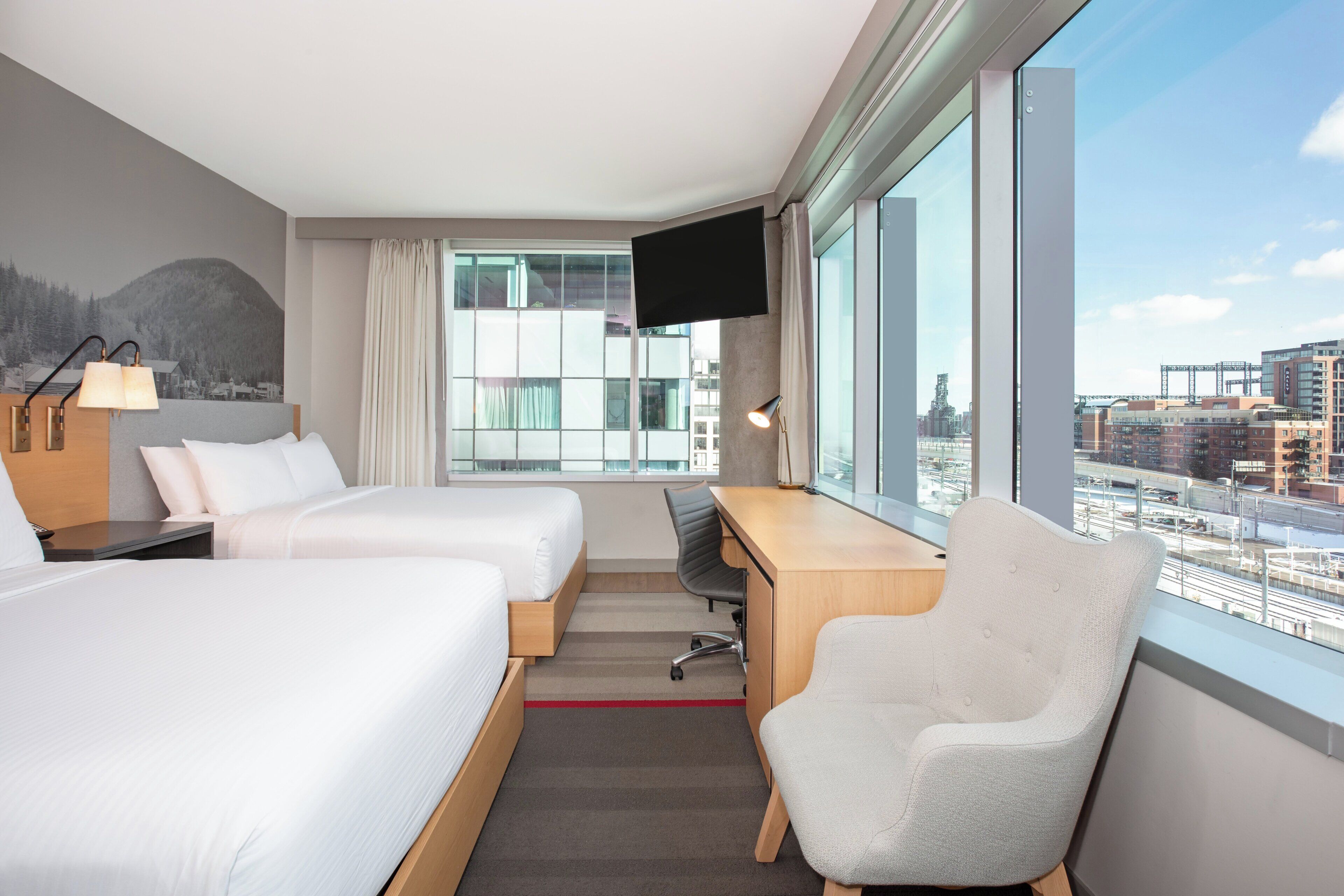 Quarto de hotel com duas camas, uma escrivaninha, uma cadeira, televisão e uma janela grande com vista para a cidade.