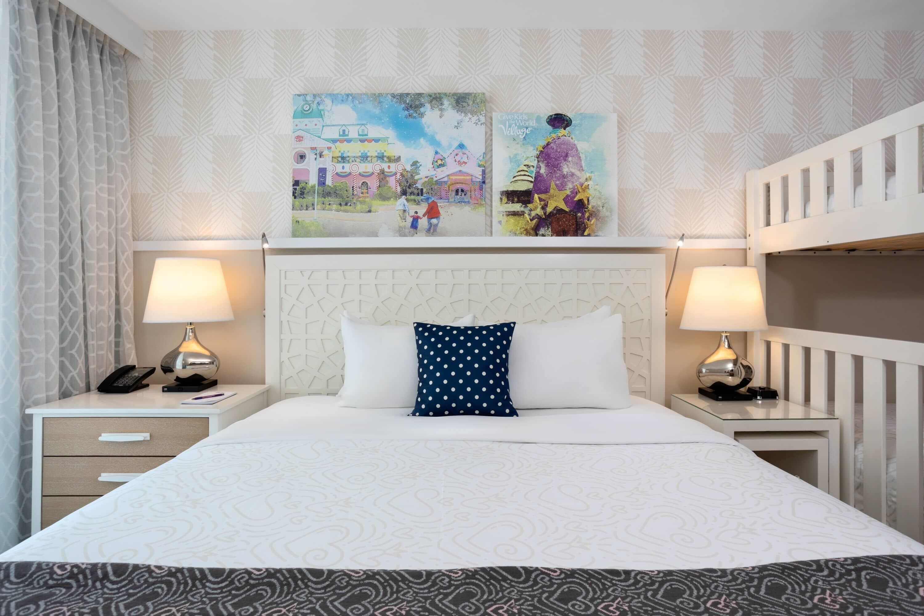 Quarto de hotel com cama, duas mesas de cabeceira com luminárias, um criado-mudo e uma cabeceira branca, além de duas obras de arte na parede.
