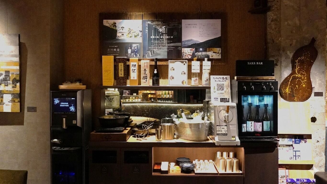 Uma área de bar com balcão, prateleiras com garrafas, uma máquina de café e uma placa indicando um bar de saquê.
