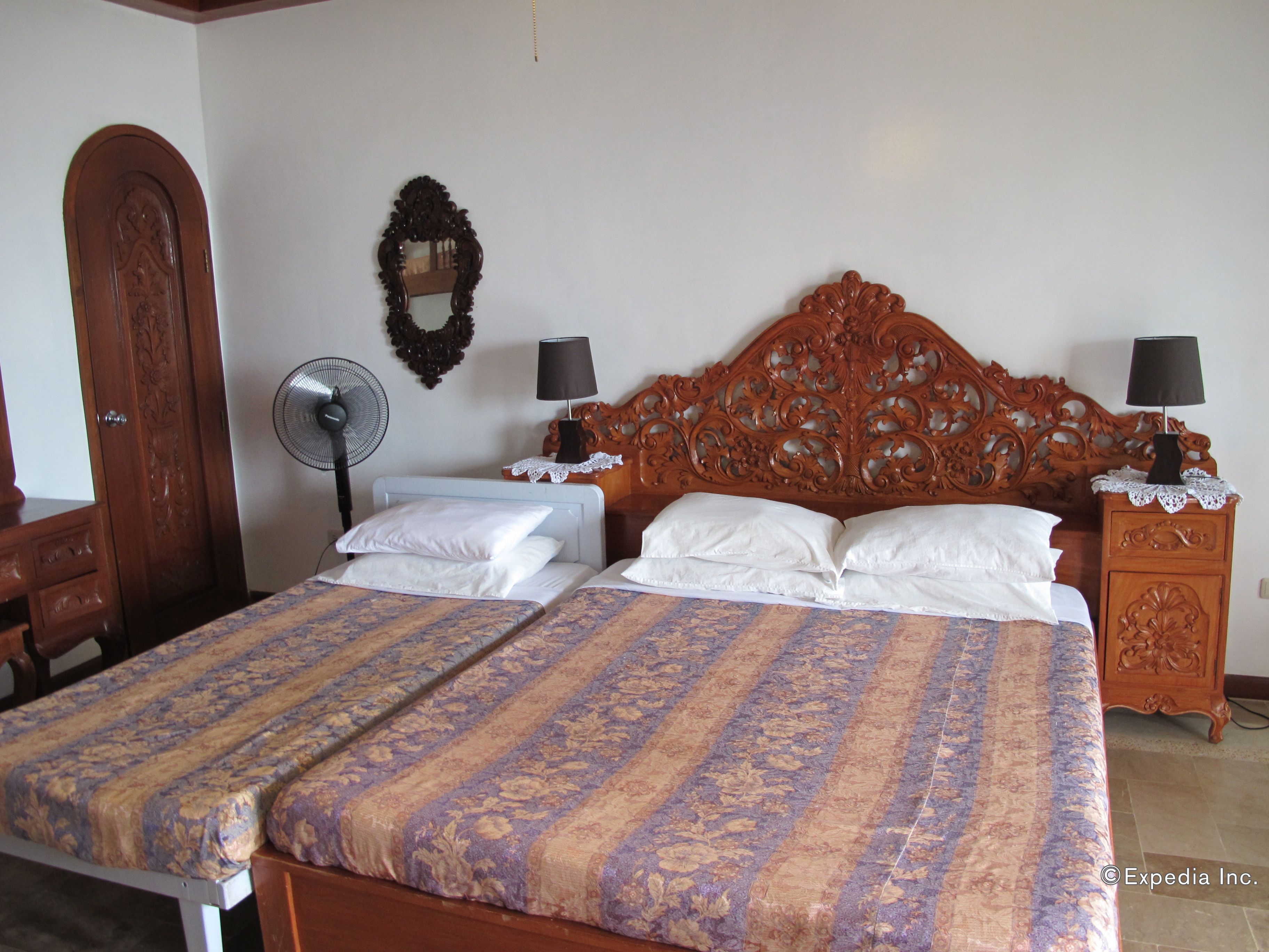 Uma cama de casal com colcha florida, cabeceira de madeira, duas mesas de cabeceira com luminárias, um ventilador fixado na parede e um guarda-roupa de madeira.