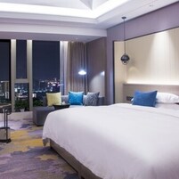 Quarto de hotel moderno com uma cama grande, área de estar com sofá e poltronas, vista para a paisagem urbana através de grandes janelas e horizonte da cidade visível à noite.