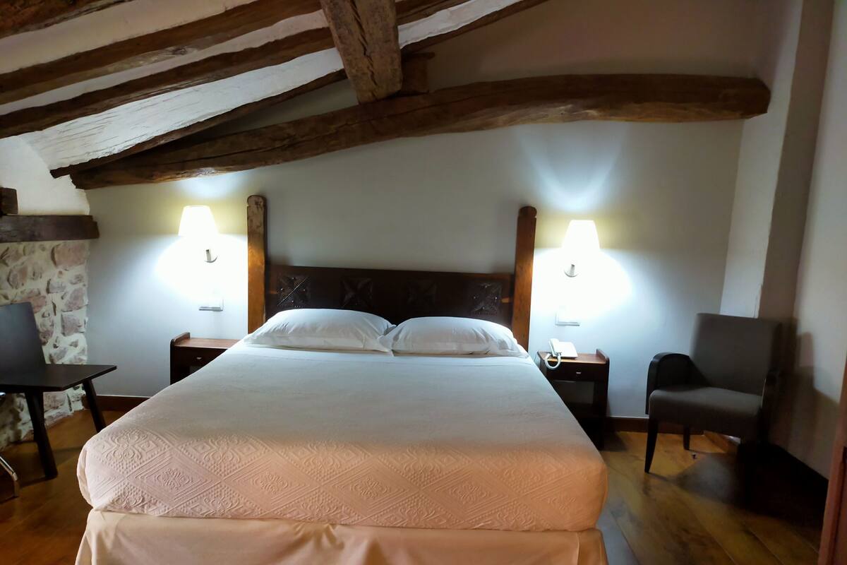 Quarto com cama grande, duas luminárias de apoio, parede de pedra, piso de madeira e uma cadeira.