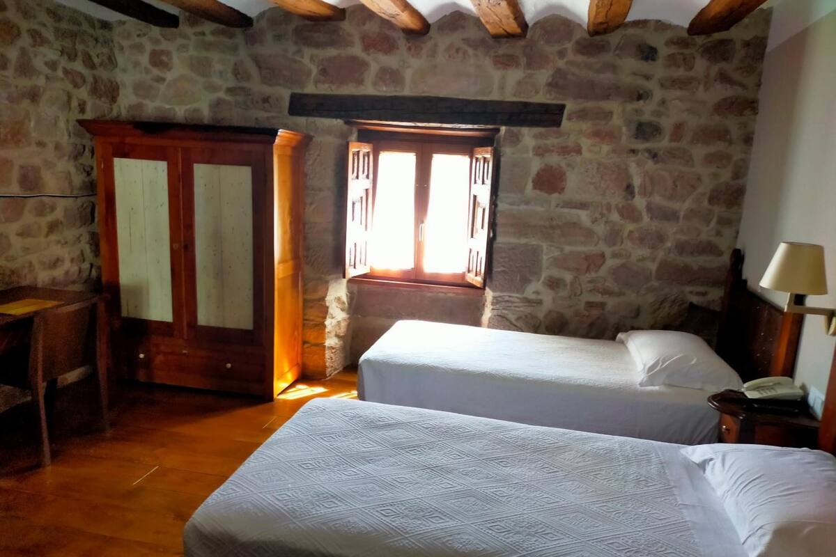 Quarto com duas camas, um guarda-roupa de madeira, uma escrivaninha e uma janela com luz solar.