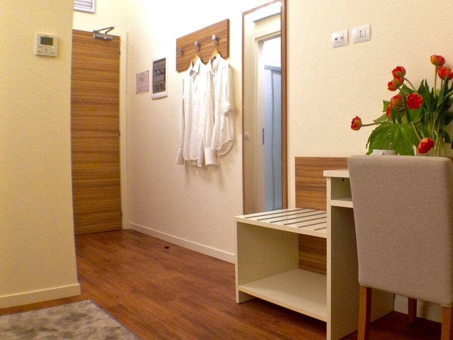 Quarto de hotel com chão de madeira, uma cadeira branca, uma pequena mesa, um espelho e um vaso com flores vermelhas.