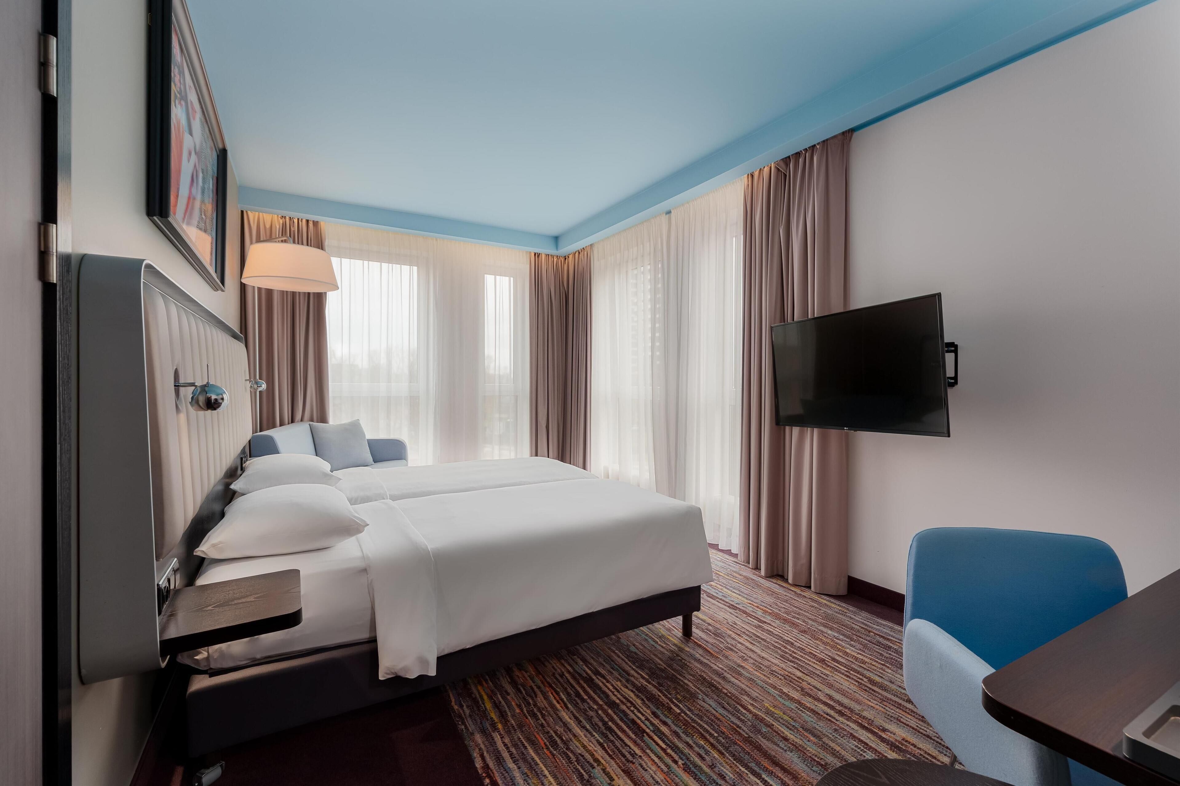 Quarto de hotel com cama, escrivaninha, televisão e janela com cortinas.