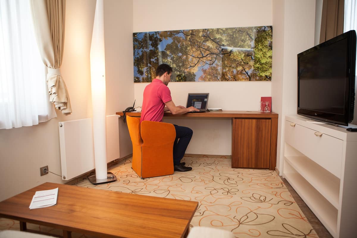 Um homem trabalhando numa mesa num quarto de hotel com uma janela grande, uma televisão de ecrã plano e um tapete estampado.