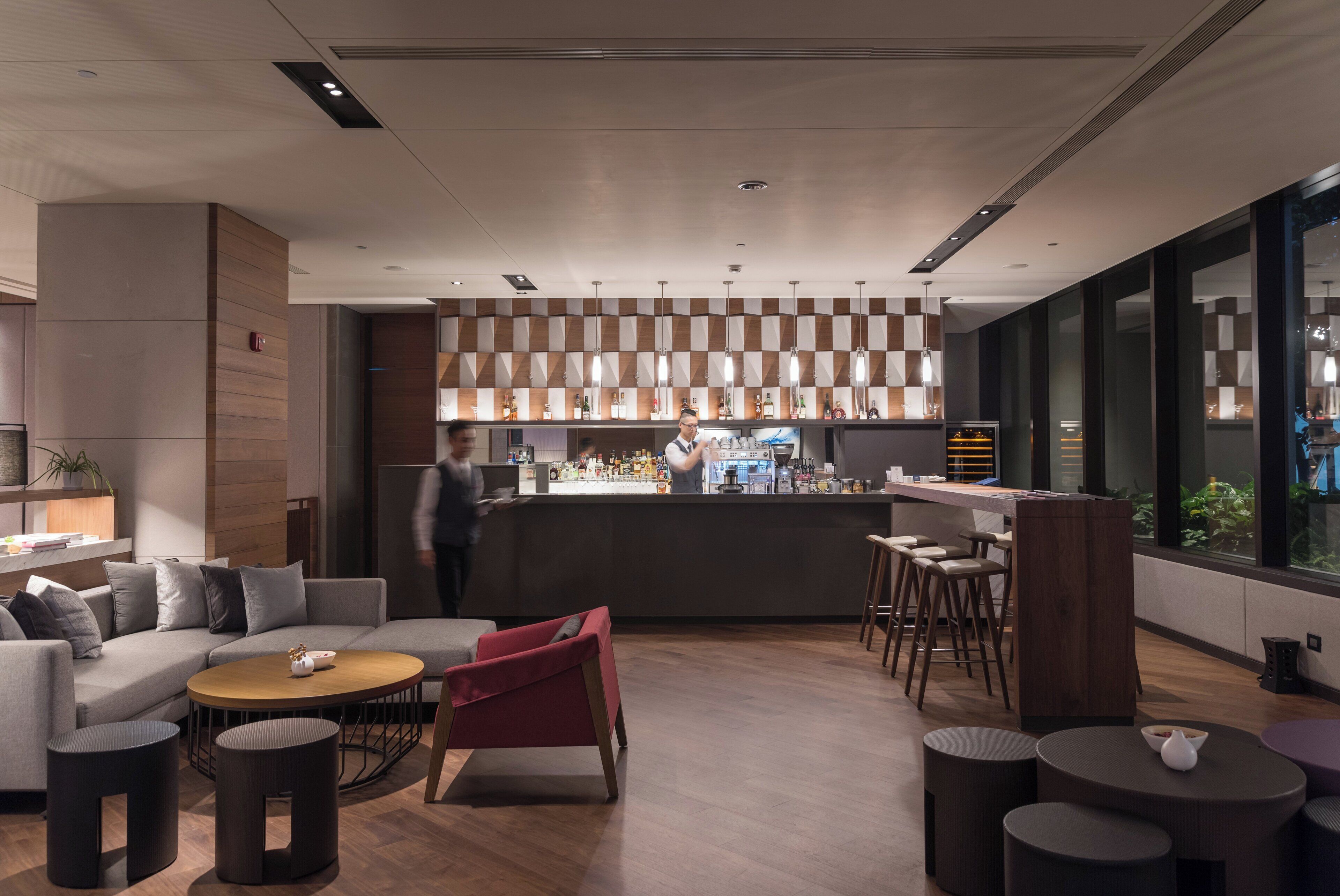 Uma área de bar moderna, com balcão, banquetas e um espaço de estar com sofás e mesas redondas.