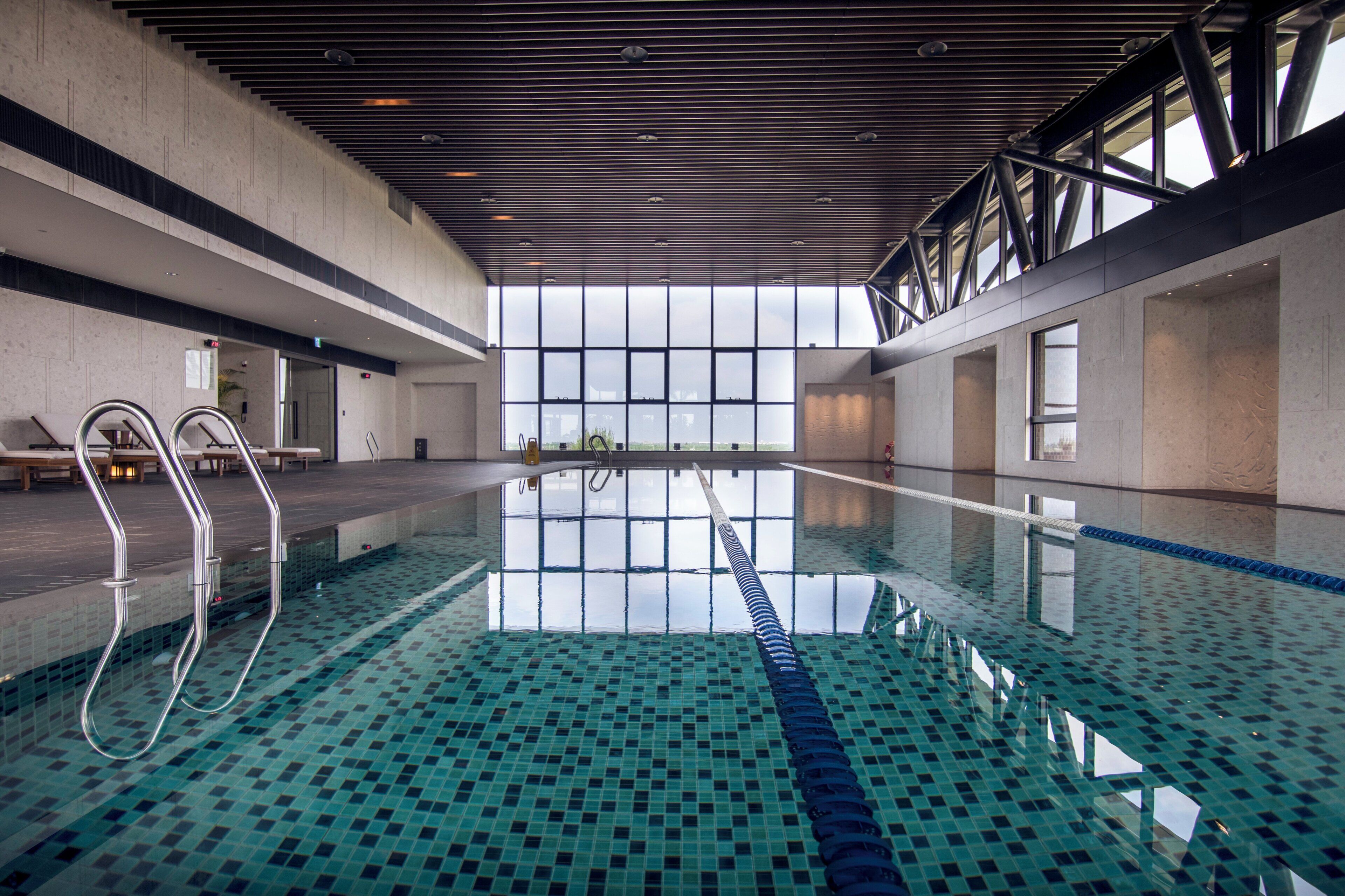Piscina interna com água cristalina, piso revestido e design arquitetônico moderno, com grandes janelas e estrutura metálica.