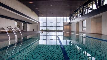 Piscina interna com água cristalina, piso revestido e design arquitetônico moderno, com grandes janelas e estrutura metálica.