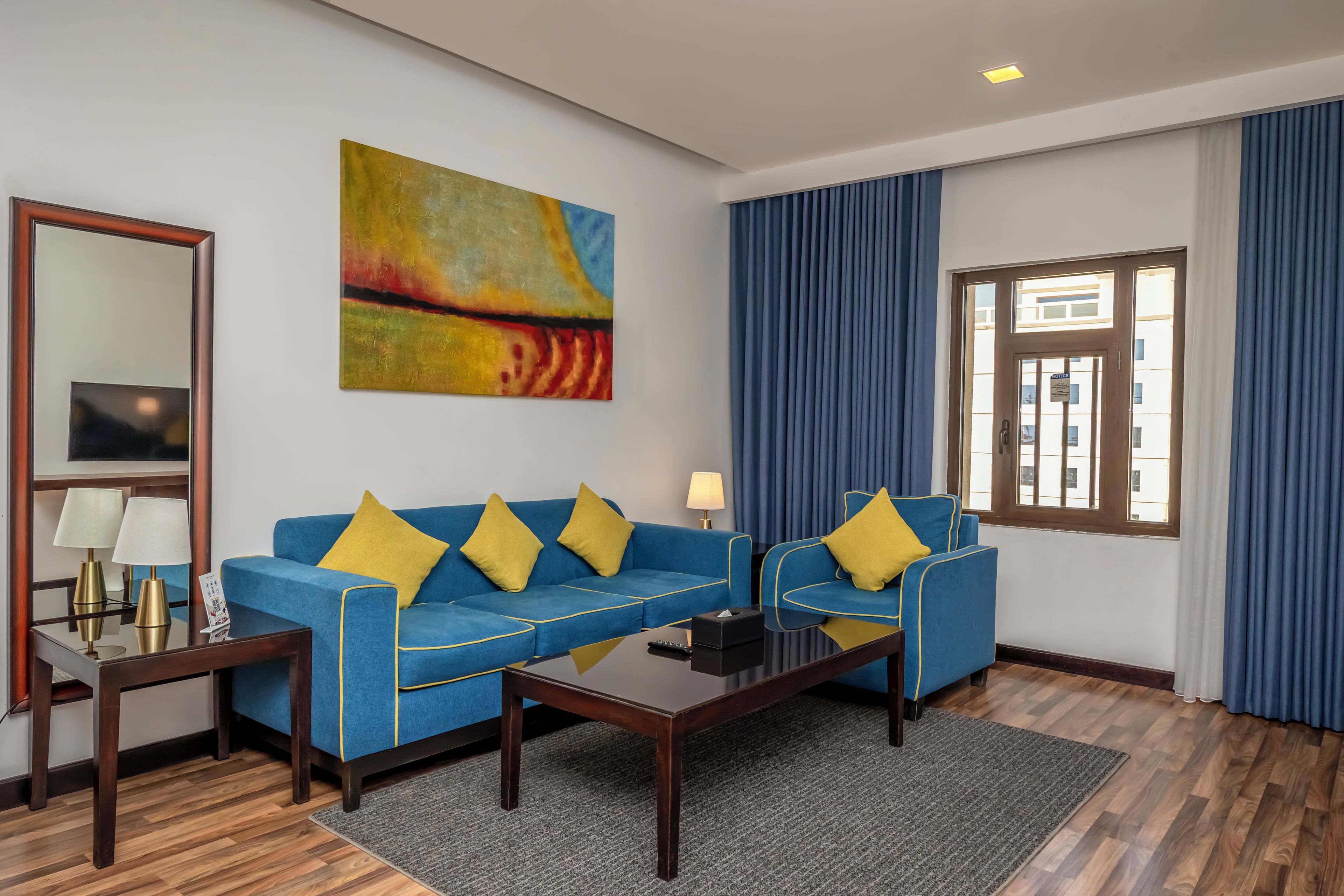 Quarto de hotel moderno com sofá azul, almofadas amarelas, mesa de centro, mesa lateral com luminárias, quadro na parede e janela com cortinas azuis.