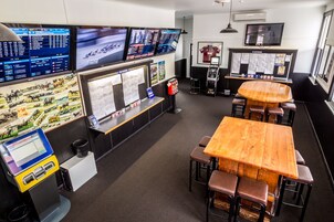 Uma sala de controle com vários monitores, uma mesa de madeira e banquetas.