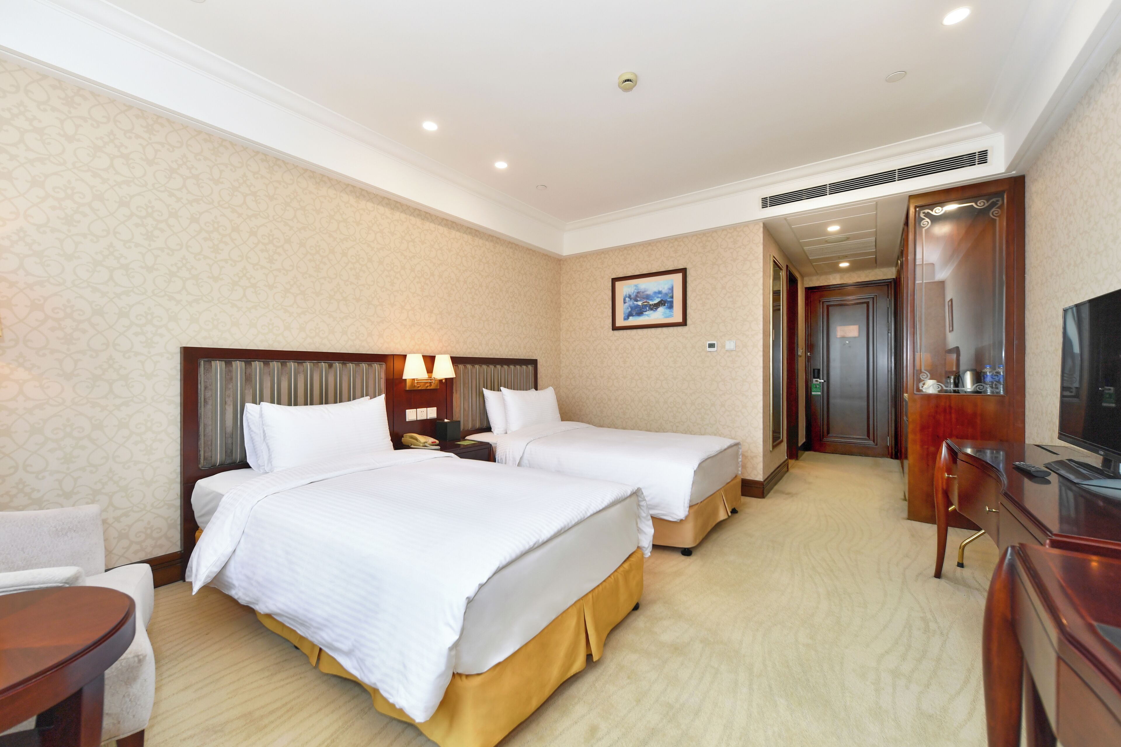 Quarto de hotel com duas camas, uma escrivaninha, uma cadeira, uma televisão e um banheiro visível por uma porta aberta.