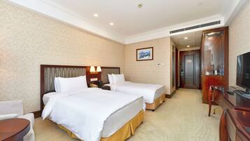 Quarto de hotel com duas camas, uma escrivaninha, uma cadeira, uma televisão e um banheiro visível por uma porta aberta.