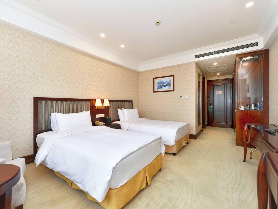 Quarto de hotel com duas camas, uma escrivaninha, uma cadeira, uma televisão e um banheiro visível por uma porta aberta.