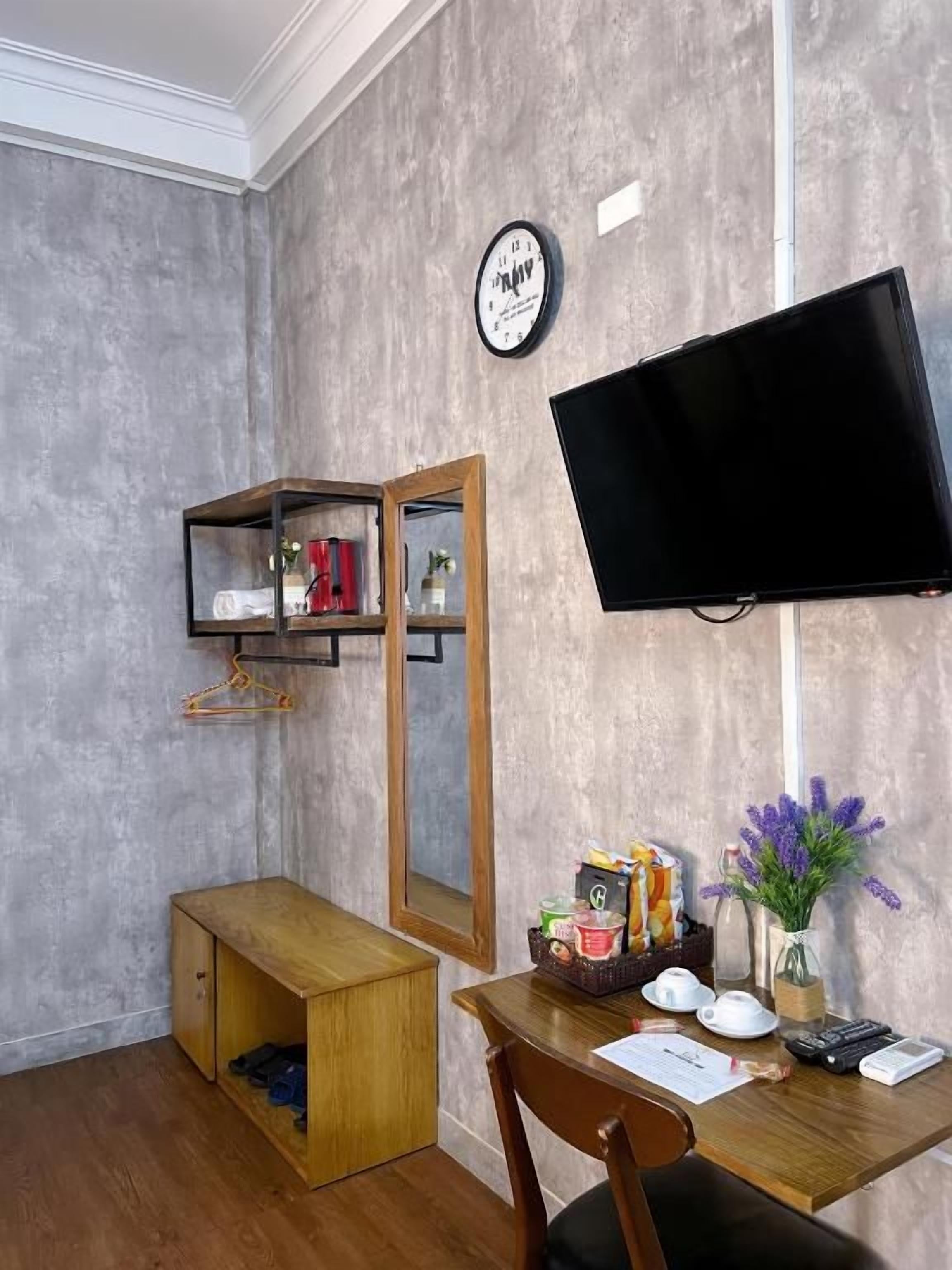 Quarto com escrivaninha de madeira, televisão de tela plana na parede, espelho e prateleira com diversos objetos.