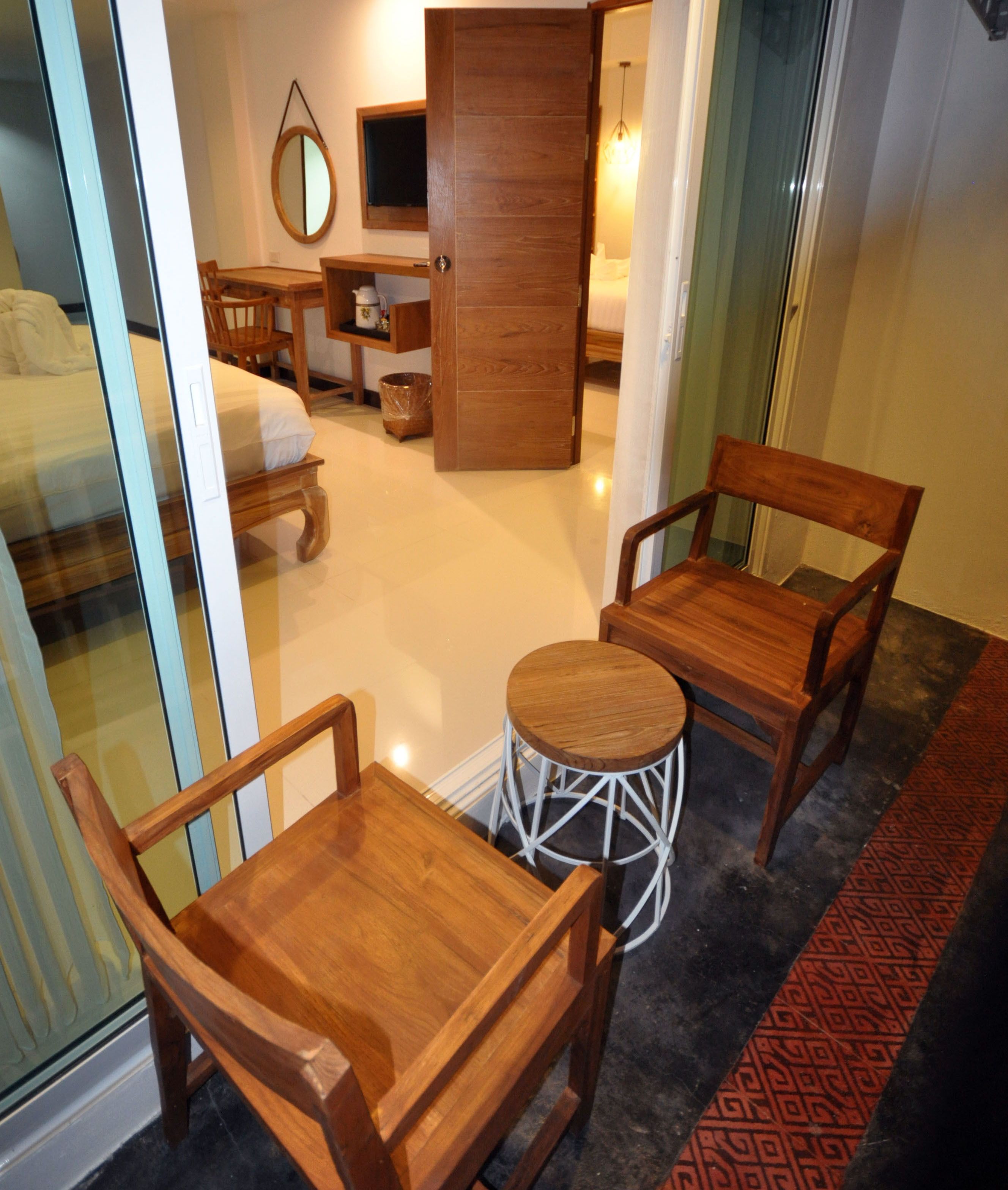 Quarto de hotel com uma mesa e cadeiras de madeira, um banquinho redondo, uma cama, uma televisão e um espelho na parede.