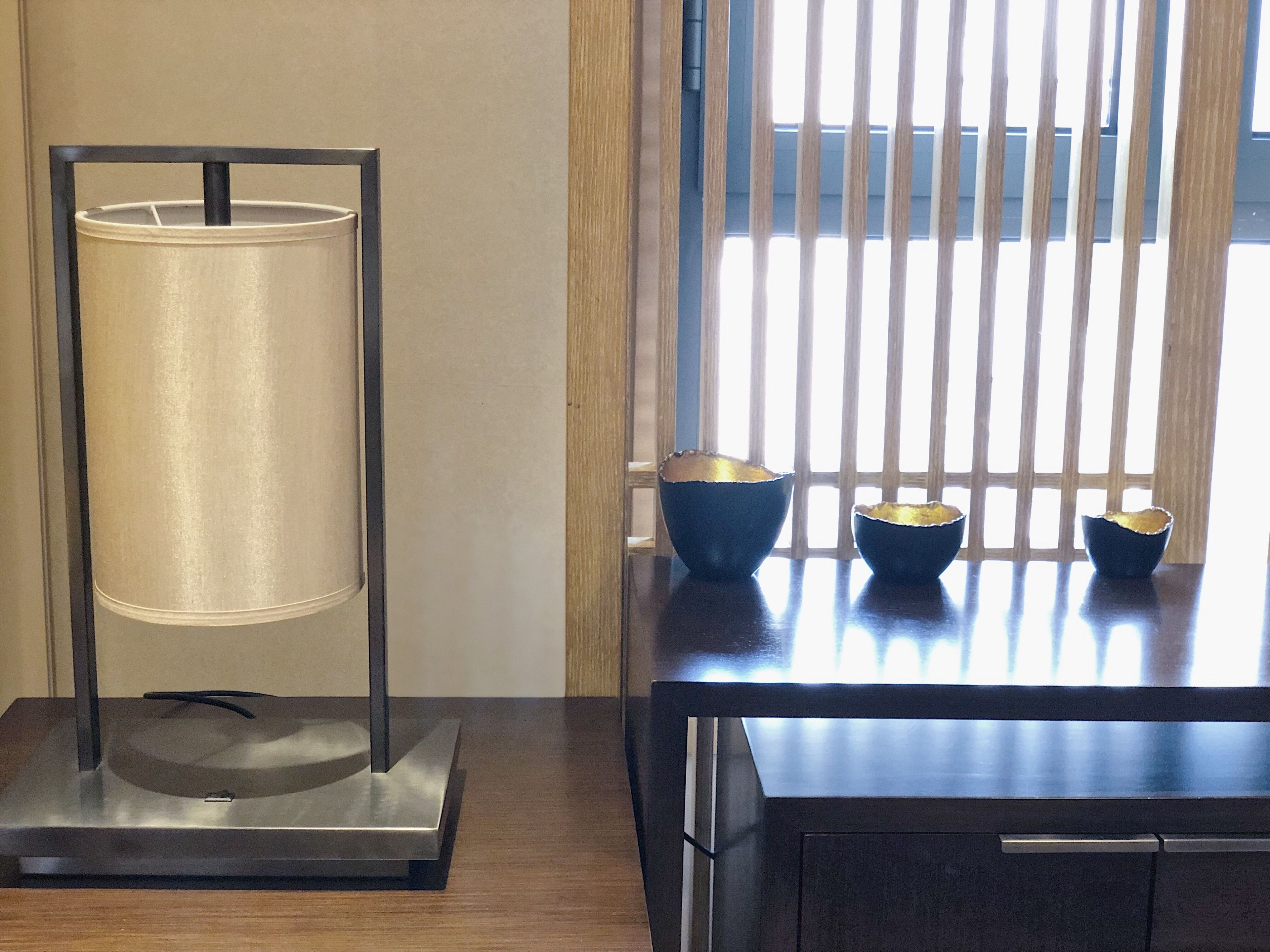 Um quarto com uma luminária sobre uma mesa, uma janela com persianas, e três recipientes no parapeito da janela.