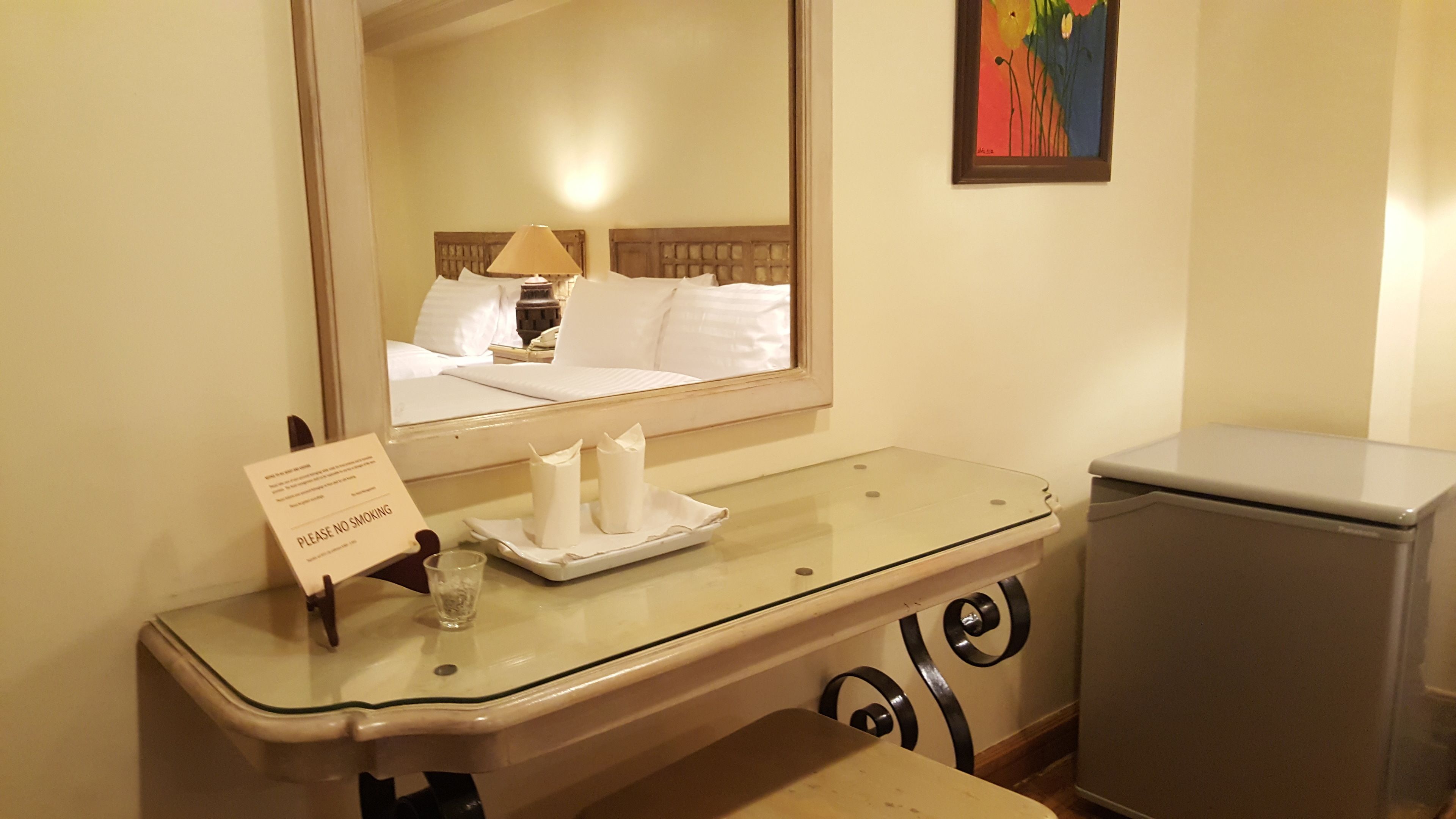Quarto de hotel com uma cama, uma mesa de cabeceira com o aviso "Não Fumar", uma pequena mesa com um copo de água, uma luminária e um frigorífico.