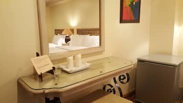 Quarto de hotel com uma cama, uma mesa de cabeceira com o aviso "Não Fumar", uma pequena mesa com um copo de água, uma luminária e um frigorífico.