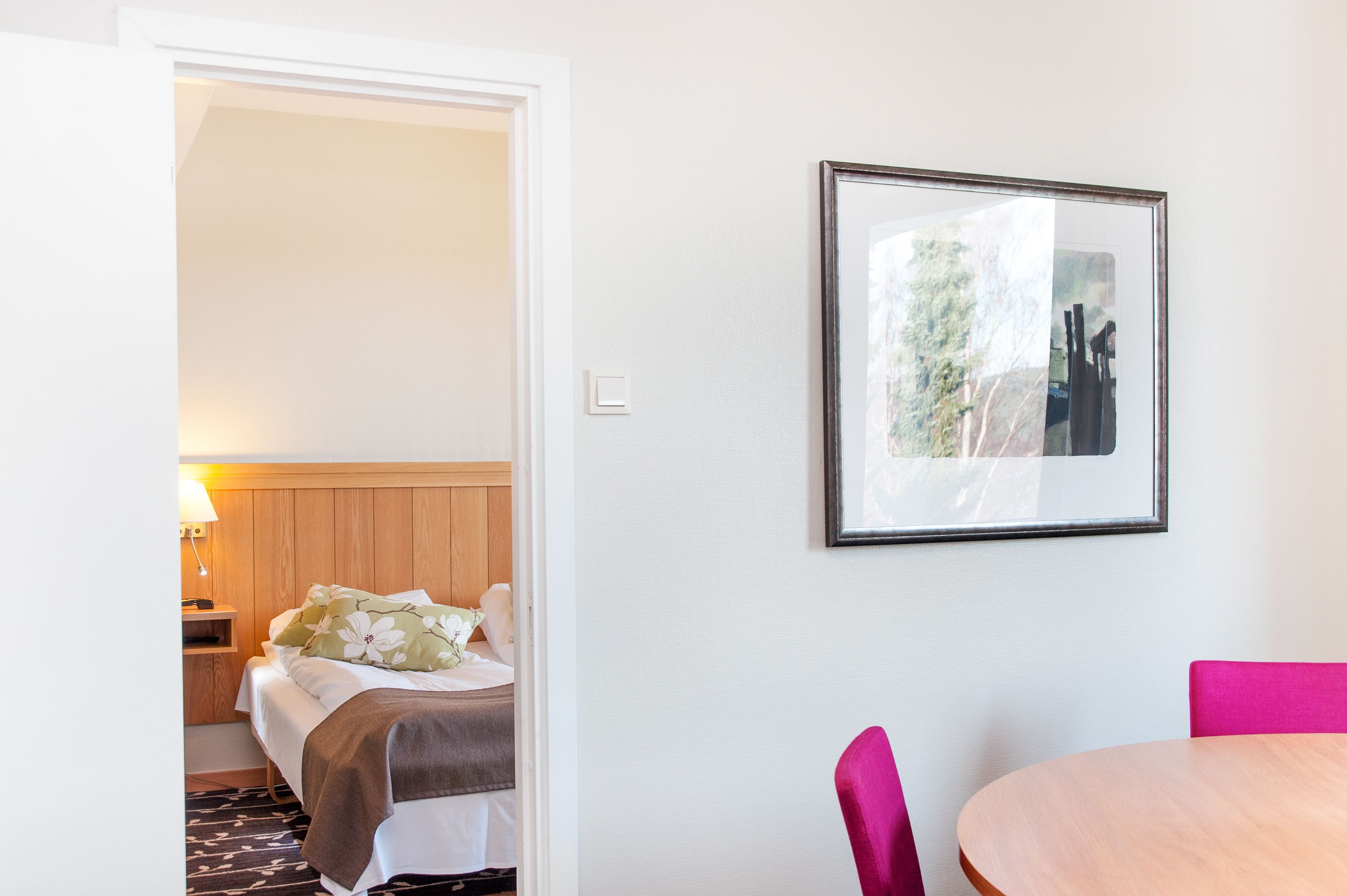 Quarto de hotel com cama, cabeceira de madeira, mesa de cabeceira com abajur, quadro na parede e mesa redonda de madeira com uma cadeira rosa.
